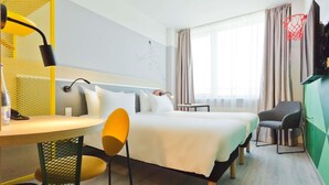 Quarto de hotel com cama, escrivaninha, cadeira, televisão e janela com cortinas.