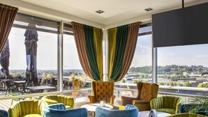 Quarto de hotel moderno com grandes janelas, varanda com área de estar e vista para a cidade.