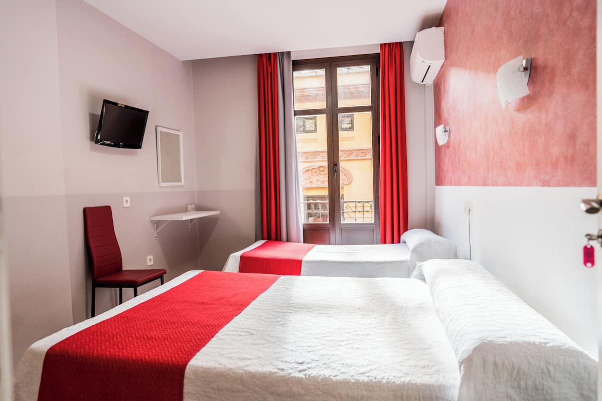 Quarto de hotel com duas camas, uma televisão, uma cadeira vermelha e uma janela com cortinas vermelhas.