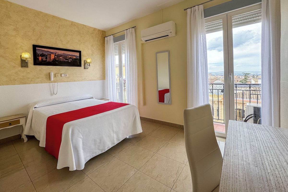 Quarto de hotel com cama, mesinha de cabeceira, cadeira, varanda e televisão na parede.
