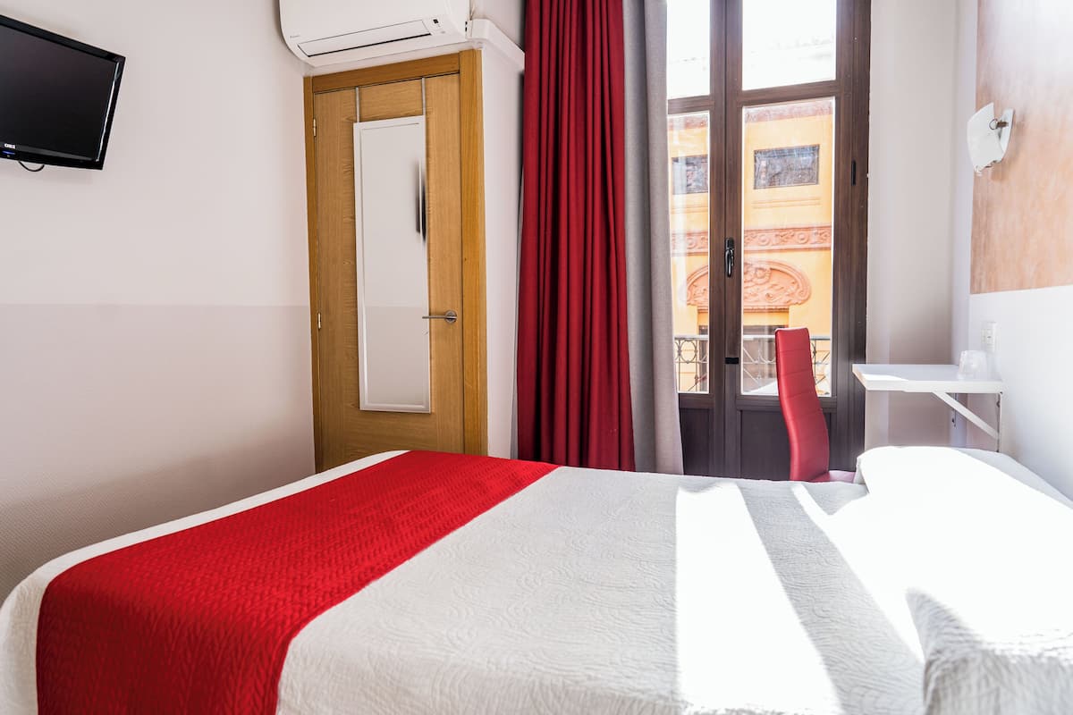 Quarto de hotel com cama, televisão, janela com cortinas vermelhas e vista para o edifício exterior.
