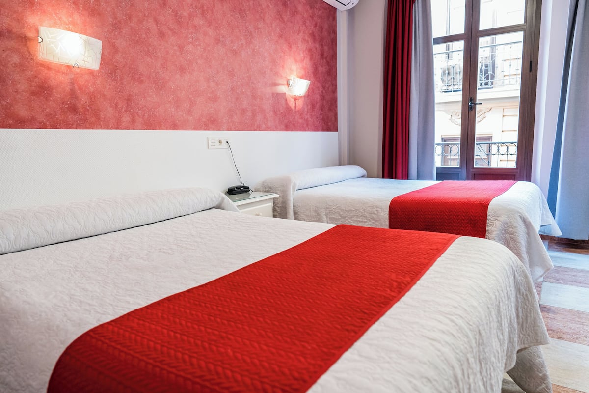 Quarto de hotel com duas camas, cada uma com colcha vermelha e branca, uma mesa de cabeceira com abajur e uma janela com cortinas vermelhas.