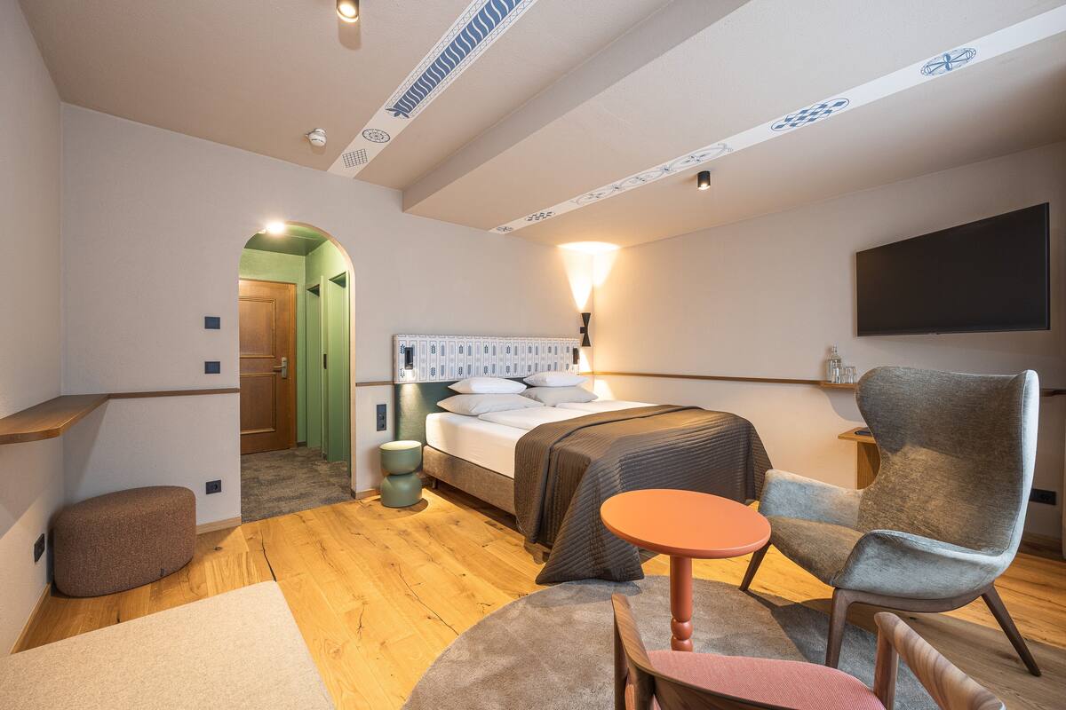 Quarto de hotel com cama, uma cadeira, uma mesa pequena e uma TV fixada na parede.
