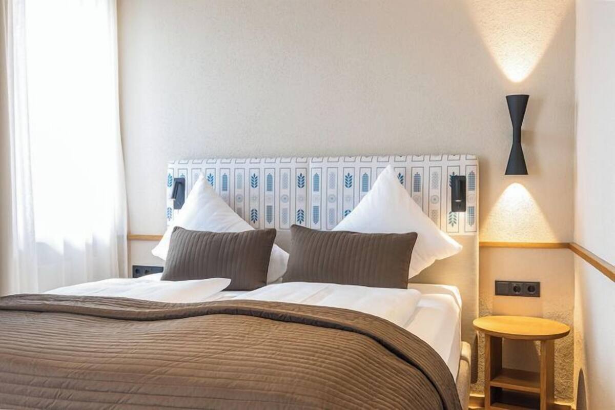 Quarto de hotel com cama grande, duas almofadas, mesa de cabeceira e luminária na parede.