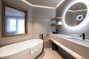 Uma casa de banho moderna com uma banheira independente, um espelho grande com luz circular e uma janela com cortinas leves.