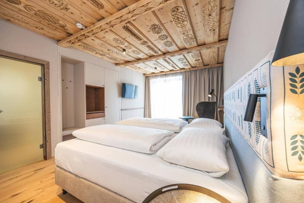 Quarto com teto de madeira, cama com roupa de cama branca, televisão na parede e janela com cortinas.