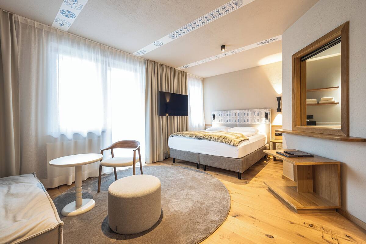 Quarto de hotel moderno com cama, escrivaninha, cadeira, um banquinho redondo e um espelho grande.