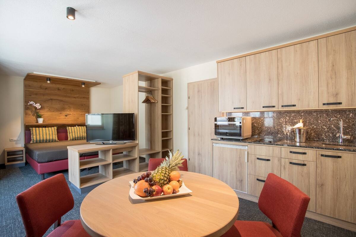Quarto de hotel moderno com mesa de jantar de madeira, cadeiras vermelhas, um sofá, uma TV, área de cozinha e uma fruteira.