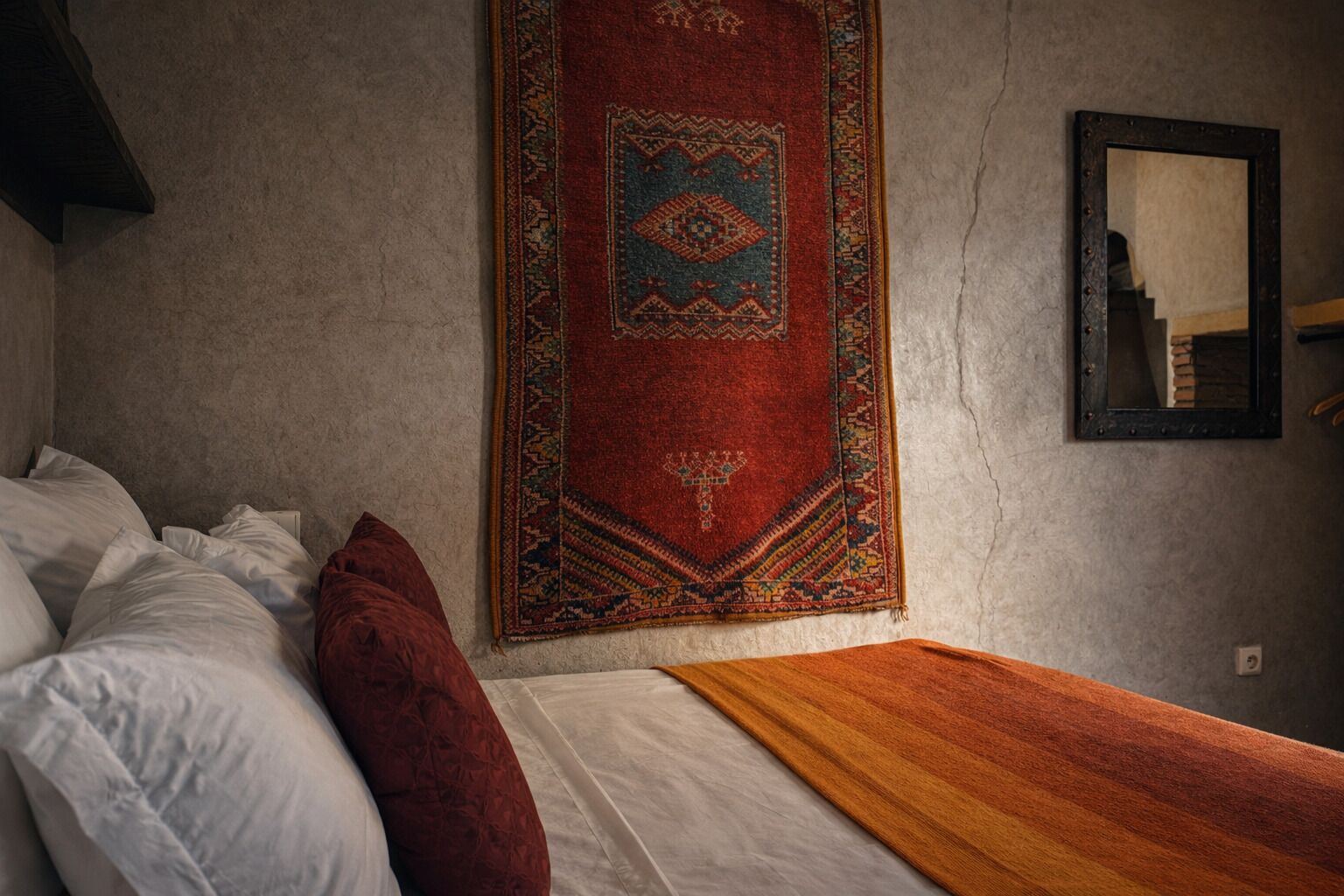 Quarto com cama, tapete estampado em vermelho e azul, cabeceira de madeira e espelho na parede.