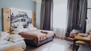 Quarto de hotel com uma cama, duas mesas de cabeceira, uma escrivaninha com cadeira e uma janela grande com cortinas.