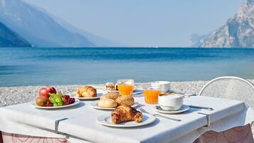 Mesa posta com itens de café da manhã, incluindo croissants, frutas e suco, com vista para uma praia e montanhas ao fundo.