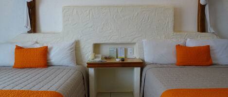 Quarto de hotel com duas camas, cada uma com colcha laranja acolchoada e travesseiros brancos. Uma pequena mesa de cabeceira de madeira com telefone, controle remoto e um cardápio está entre as camas.