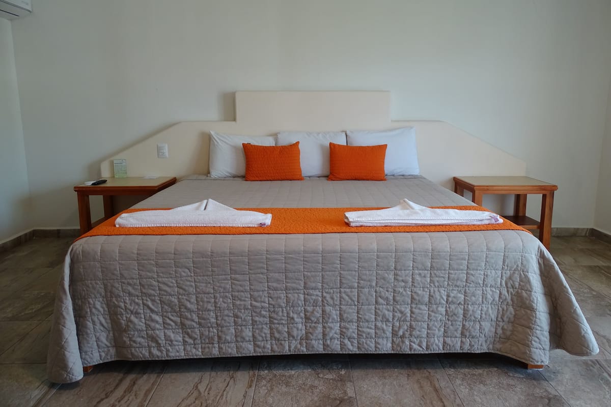 Cama bem arrumada com um edredom cinza, dois travesseiros laranja e duas toalhas brancas dobradas. Há duas mesas de cabeceira de madeira com uma pequena mesa em cada uma, e um ar condicionado branco na parede.
