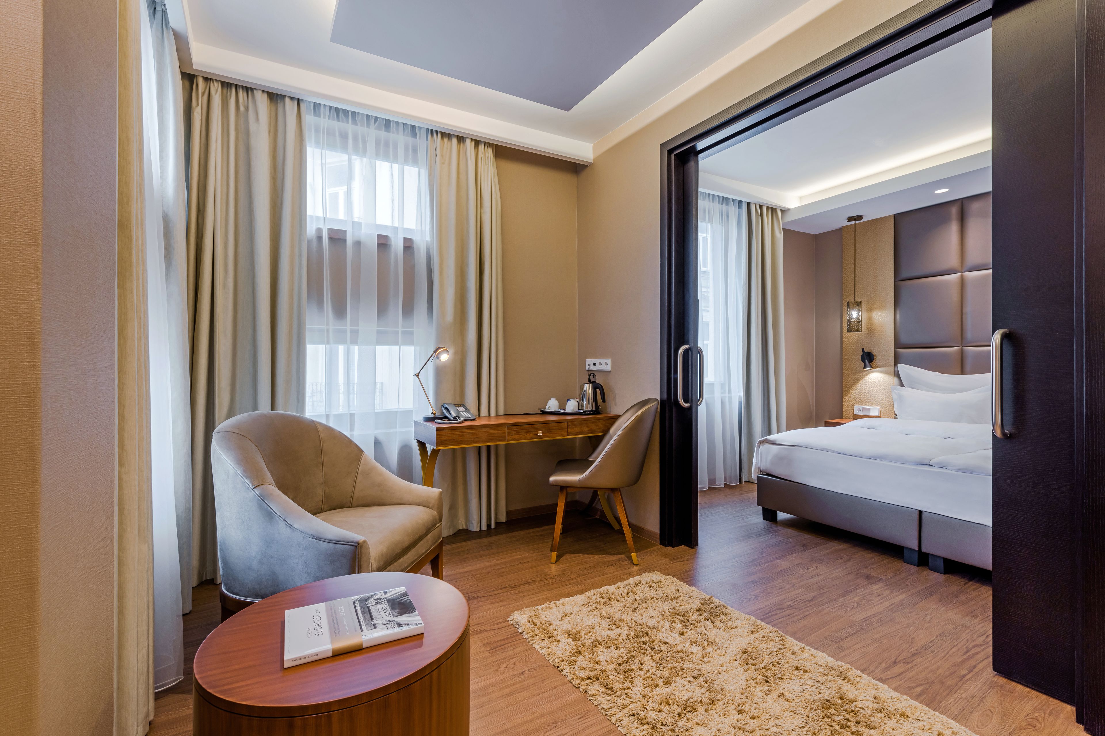 Quarto de hotel com cama, escrivaninha, cadeira, mesa redonda e uma janela grande com cortinas.