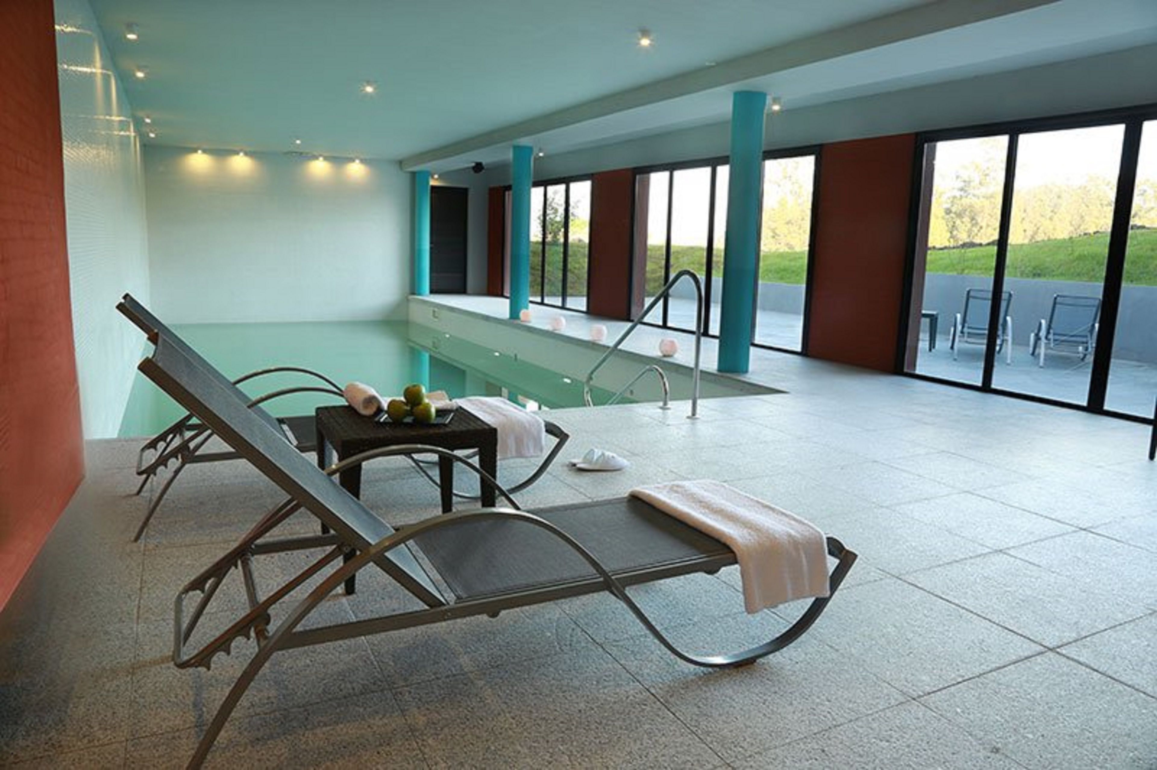 Uma área interna de piscina moderna, com cadeiras de descanso, uma mesa com frutas e grandes janelas que oferecem vista para o exterior.