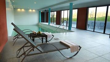 Uma área interna de piscina moderna, com cadeiras de descanso, uma mesa com frutas e grandes janelas que oferecem vista para o exterior.