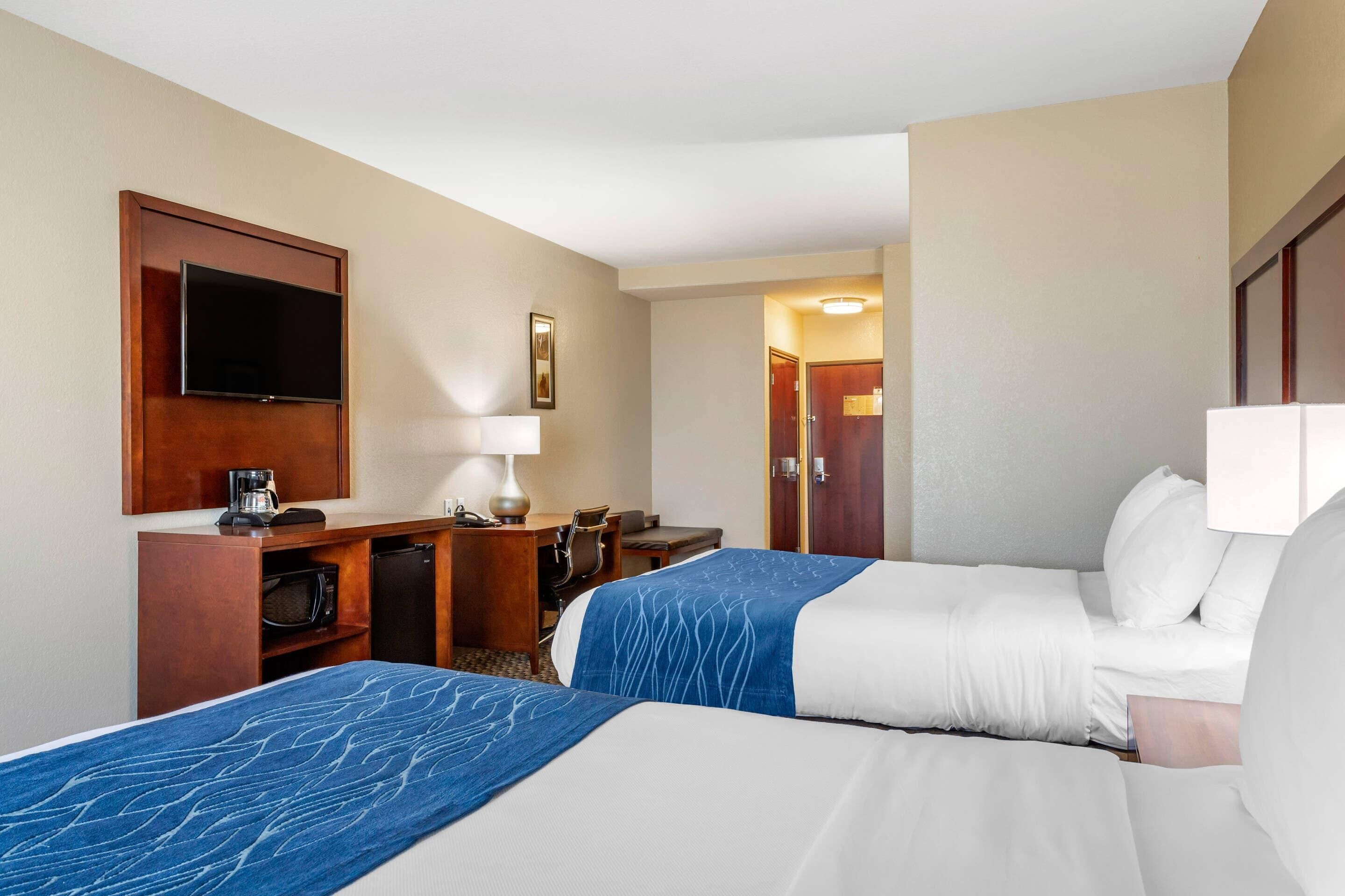 Quarto de hotel com duas camas, uma escrivaninha com televisão de tela plana, cafeteira e abajur.