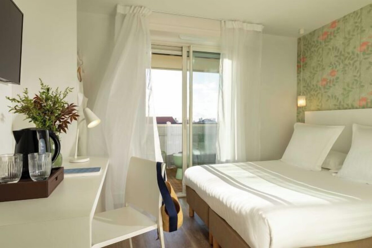 Quarto de hotel com cama, escrivaninha, cadeira, planta e janela com cortinas.