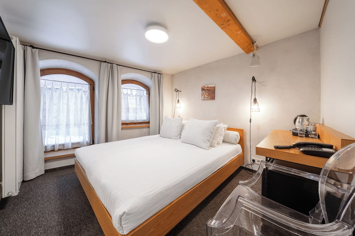 Quarto de hotel com cama, duas janelas com cortinas, escrivaninha com telefone e cafeteira.