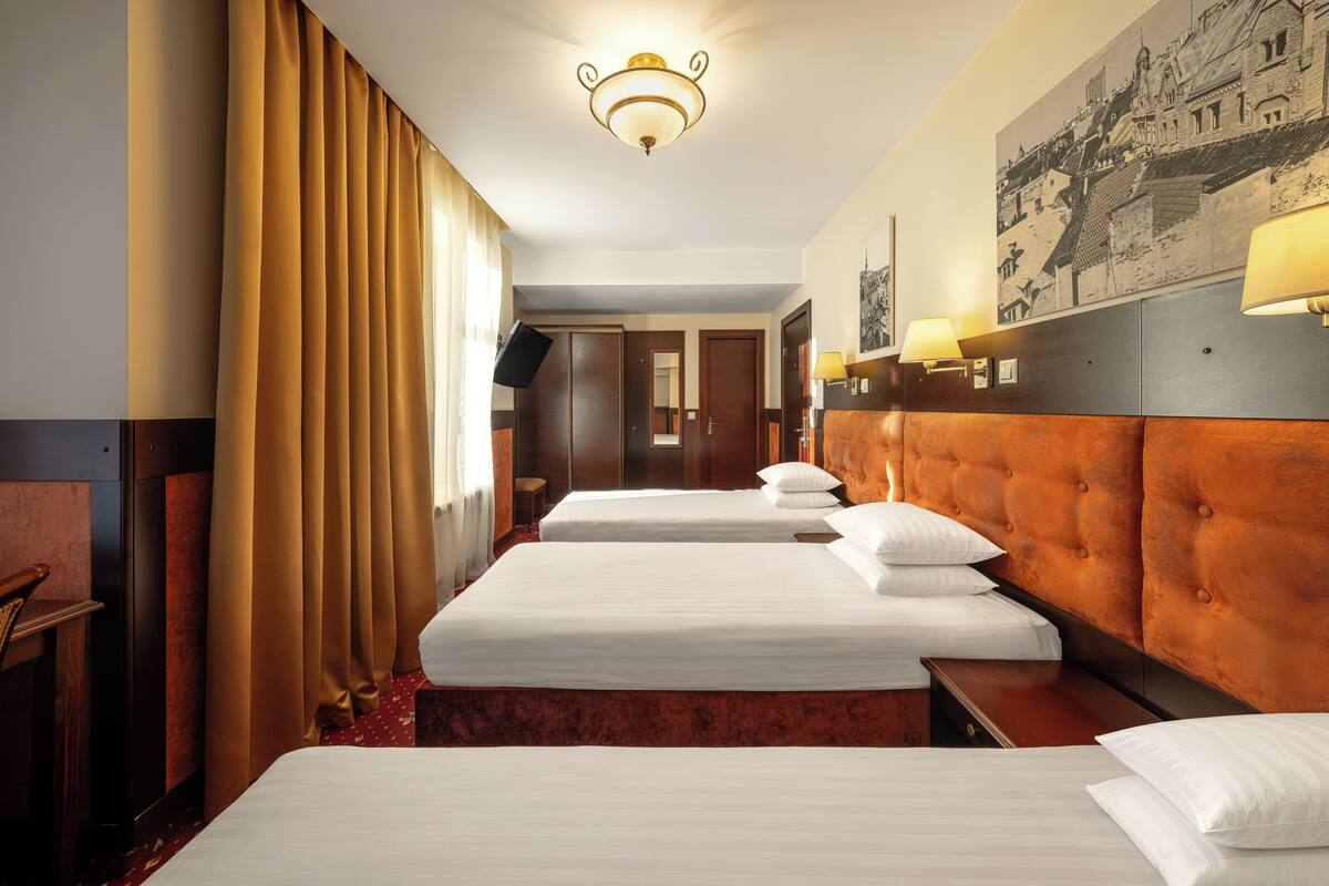 Quarto de hotel com três camas, uma janela grande com cortinas, uma televisão montada na parede e cabeceira com luminária.