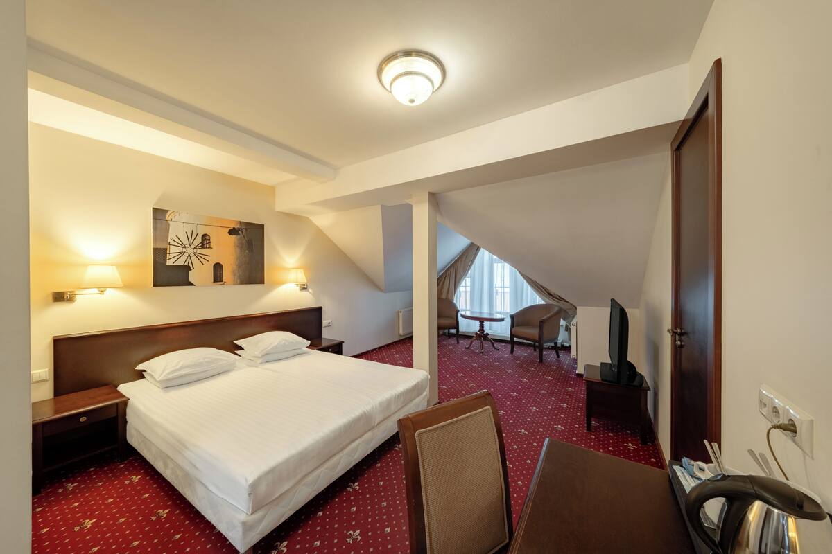 Quarto de hotel com cama, duas luminárias de cabeceira, escrivaninha com cadeira, televisão e uma mesinha.