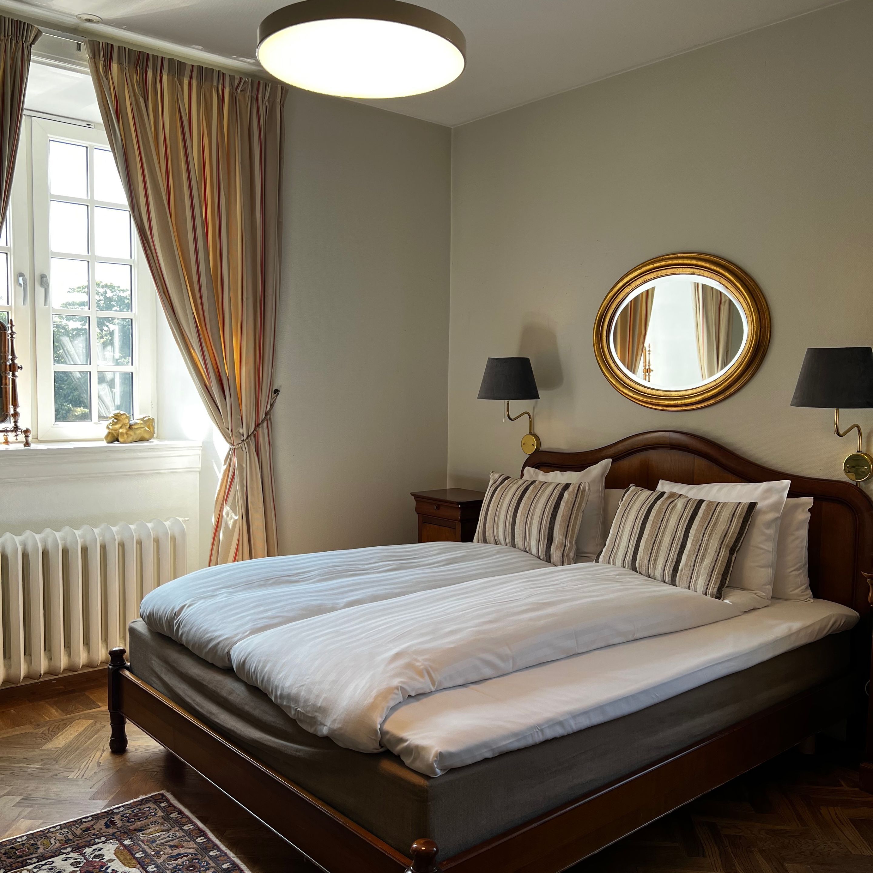 Cama bem arrumada com almofadas listradas, espelho redondo com moldura dourada e duas luminárias fixadas na parede.