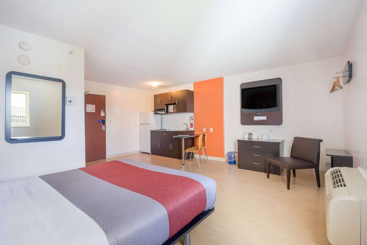 Quarto de hotel com cama, escrivaninha, cadeira, televisão e cozinha compacta.