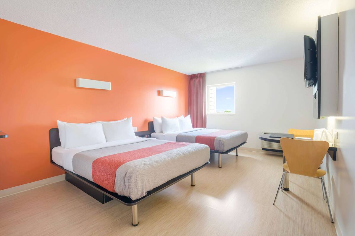 Quarto de hotel com duas camas, uma escrivaninha, uma cadeira e uma televisão na parede.
