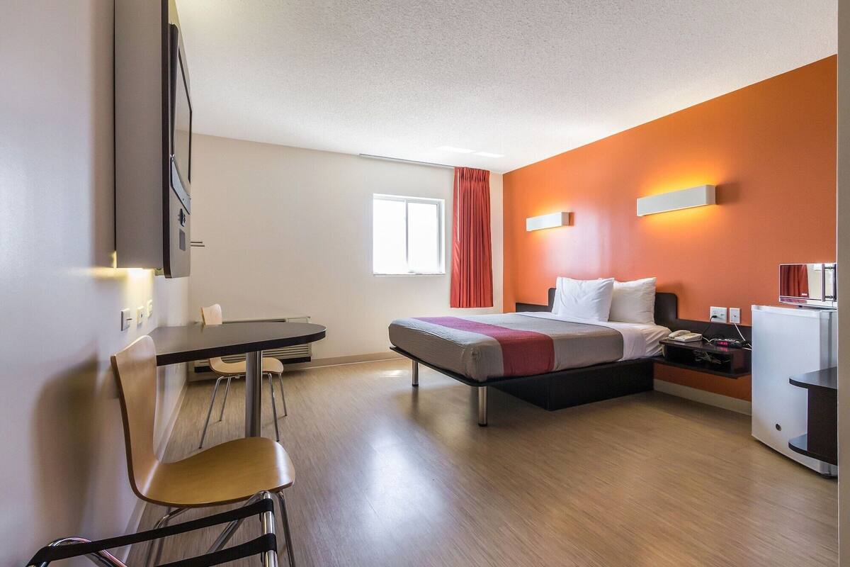 Quarto de hotel com cama, mesa de cabeceira, escrivaninha, cadeira e janela com cortinas.