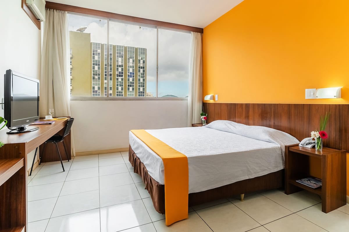 Quarto de hotel com cama, escrivaninha com televisão, mesa de cabeceira com abajur e janela com cortinas.