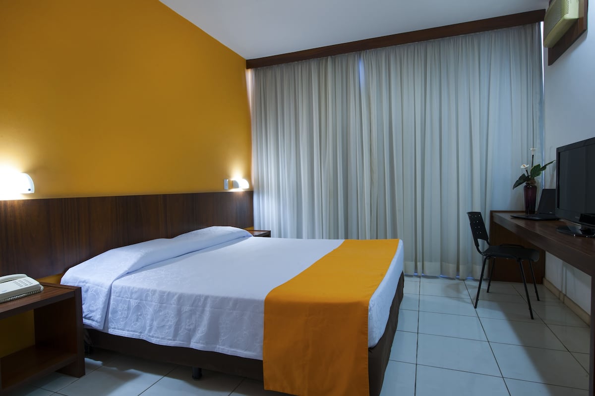 Quarto de hotel com cama, escrivaninha, cadeira, televisão e planta.