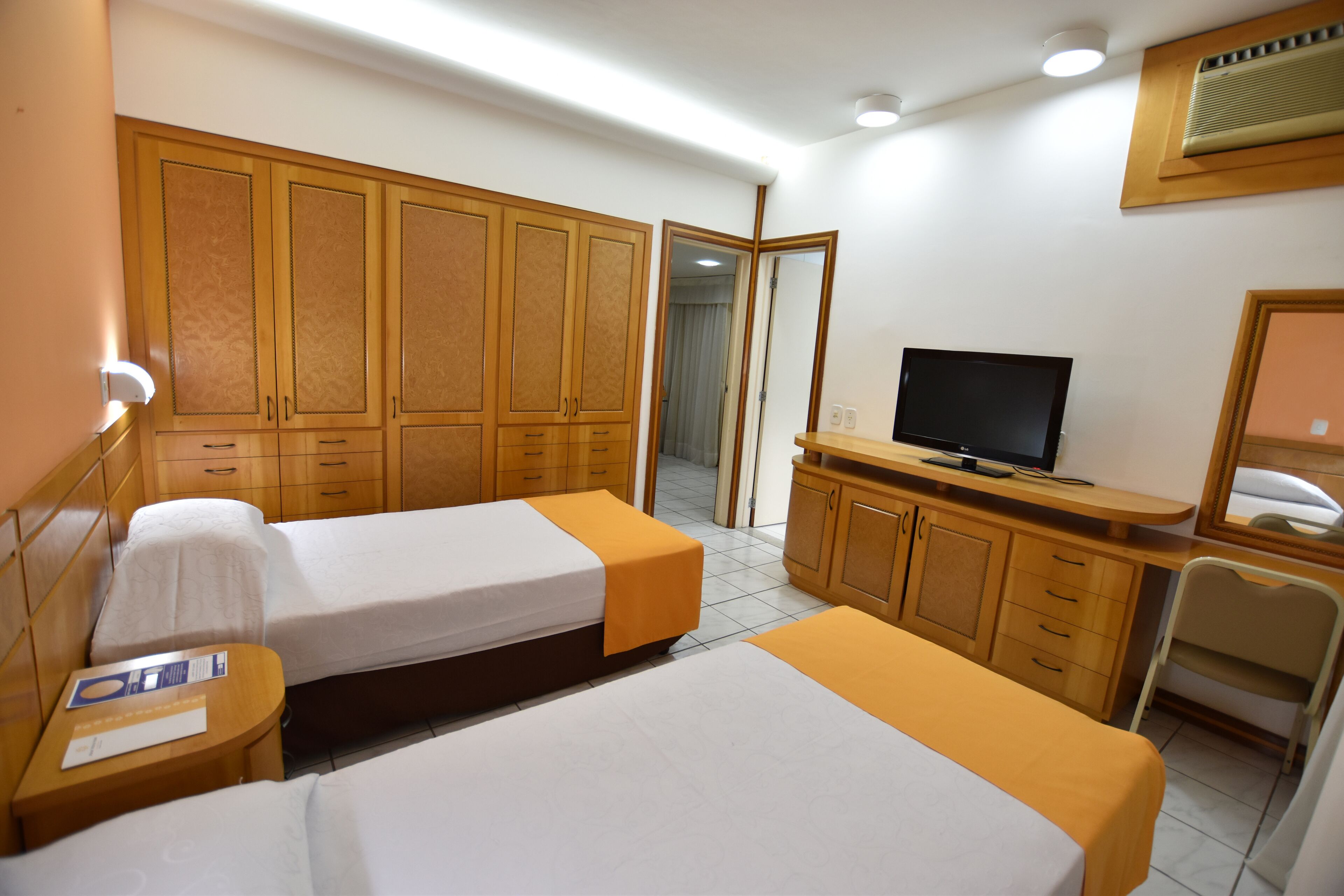 Quarto de hotel com duas camas, armários de madeira, uma televisão e uma escrivaninha.