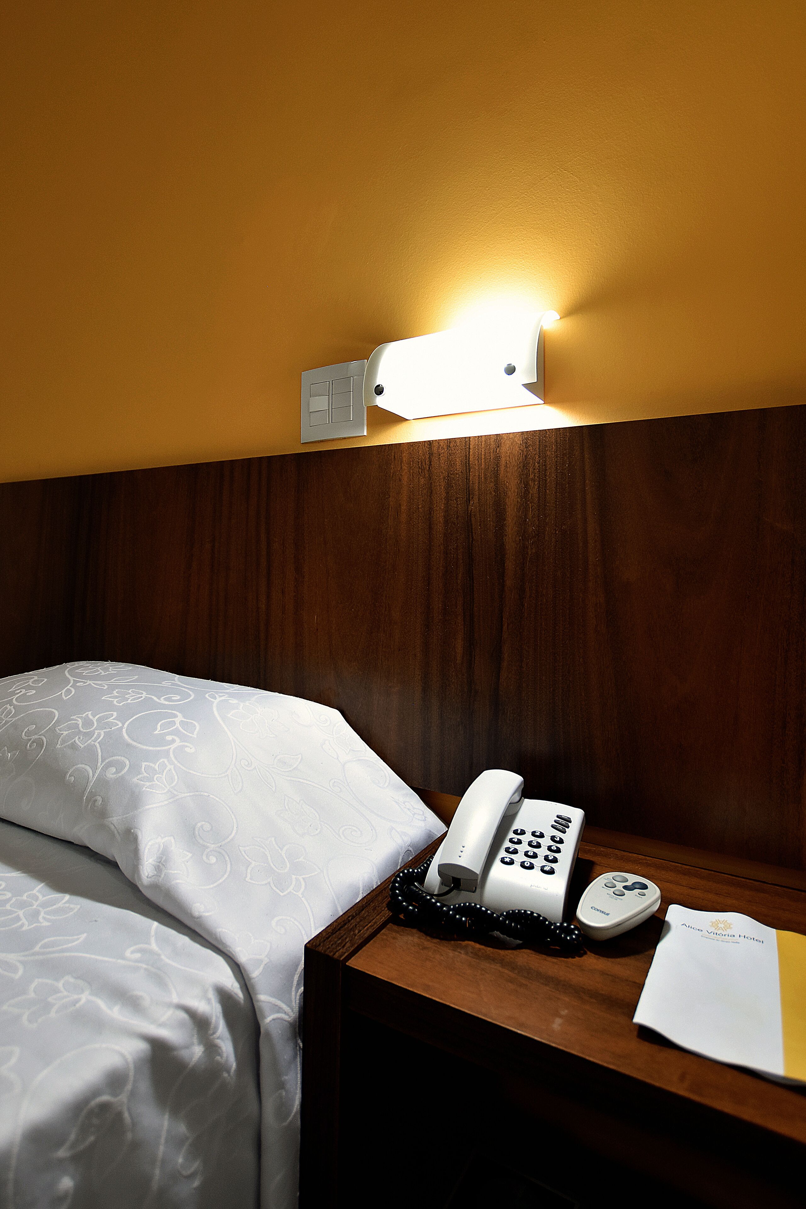 Quarto de hotel com cabeceira de madeira, mesa de cabeceira com telefone, controle remoto e um pedaço de papel.