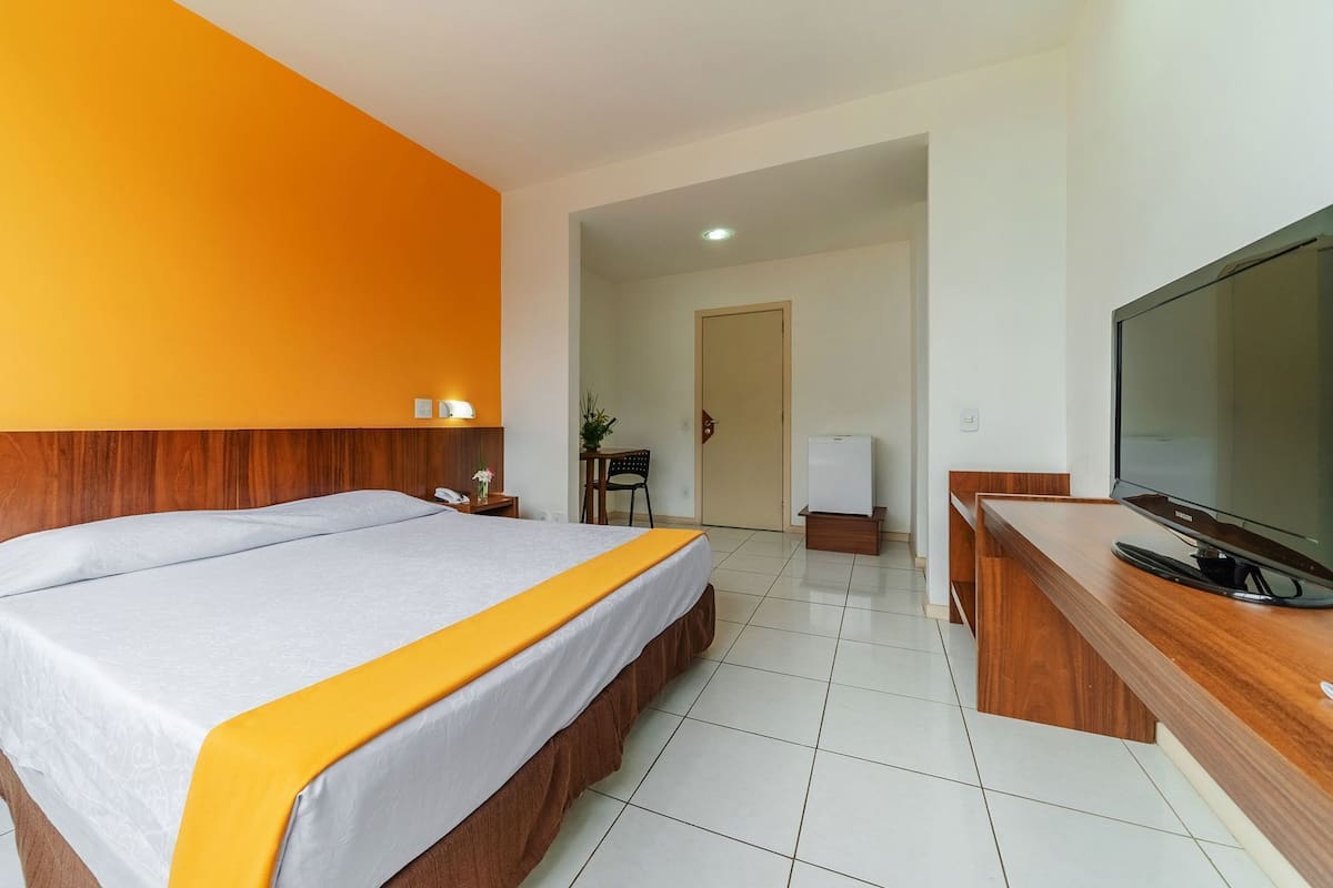Quarto de hotel com uma cama, uma escrivaninha, uma televisão e uma porta que dá para outro espaço.