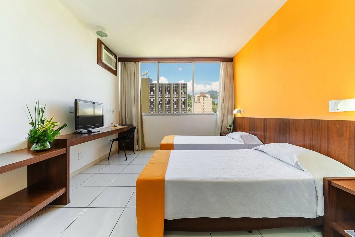 Quarto de hotel com duas camas, uma mesa de trabalho com televisão, uma planta em vaso e vista para edifícios.