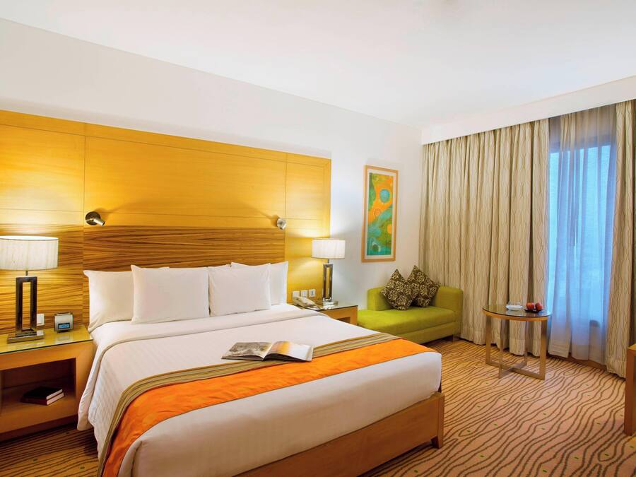 Quarto de hotel com uma cama grande, duas mesas de cabeceira com luminárias, uma poltrona verde, uma pequena mesa com uma planta e uma janela com cortinas.