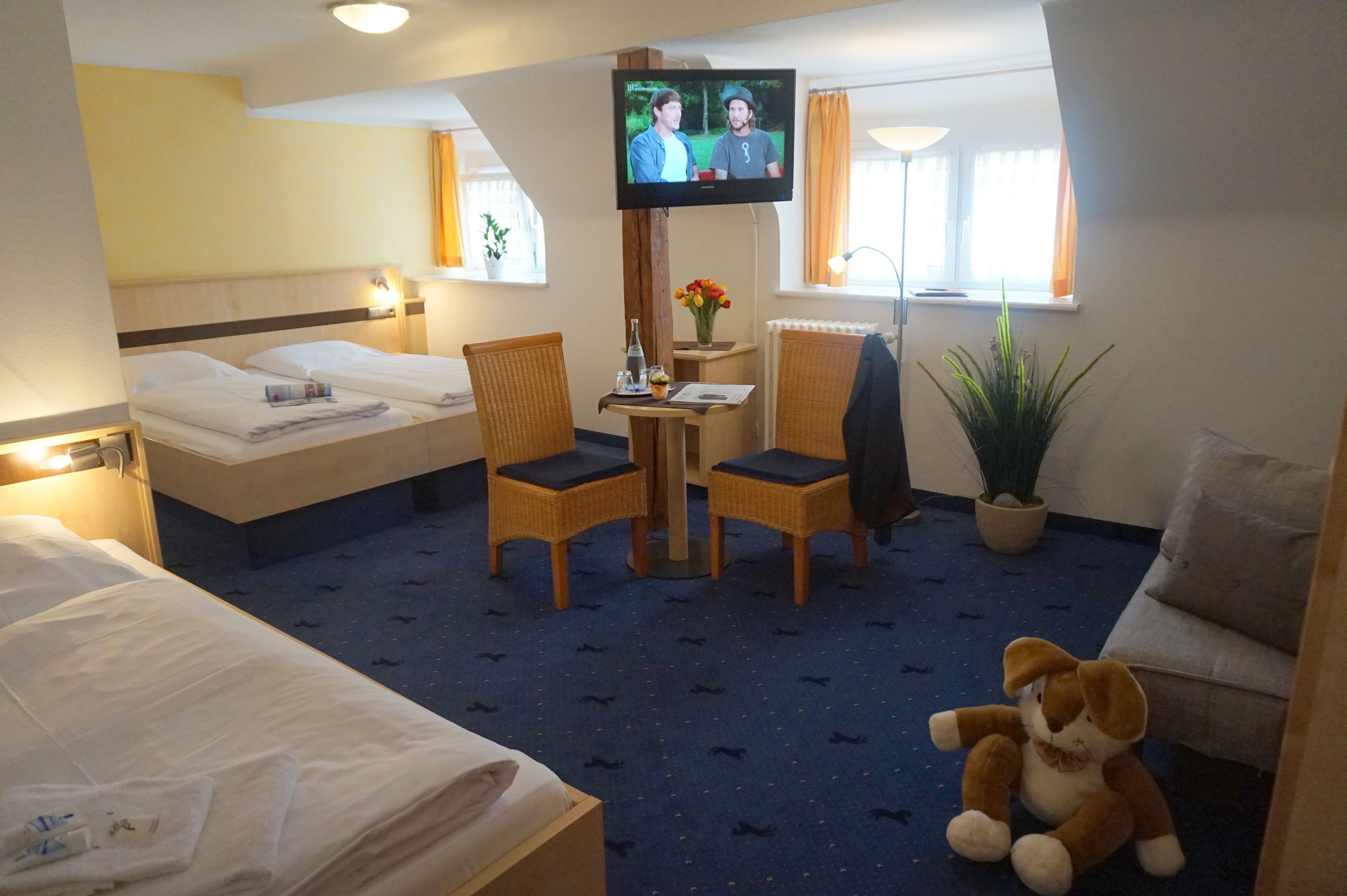 Quarto de hotel com duas camas, uma televisão, uma pequena mesa com cadeiras, um vaso de planta e um bicho de pelúcia.