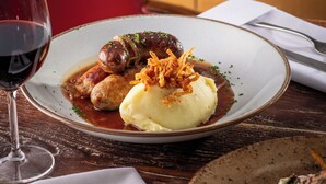Prato de comida com salsichas, purê de batatas e um copo de vinho tinto sobre uma mesa de madeira.