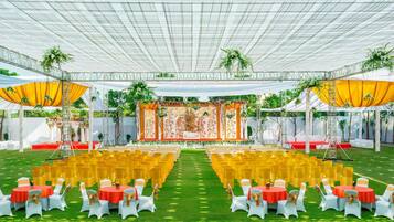 Um espaço para eventos ao ar livre com palco, fileiras de cadeiras amarelas e mesas brancas com toalhas vermelhas.