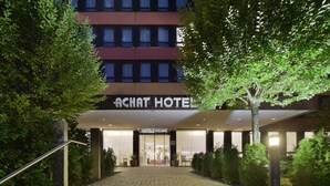 A entrada do Hotel Achat com design moderno, apresentando fachada de vidro, escada que leva à entrada e vegetação bem cuidada ao redor do prédio.