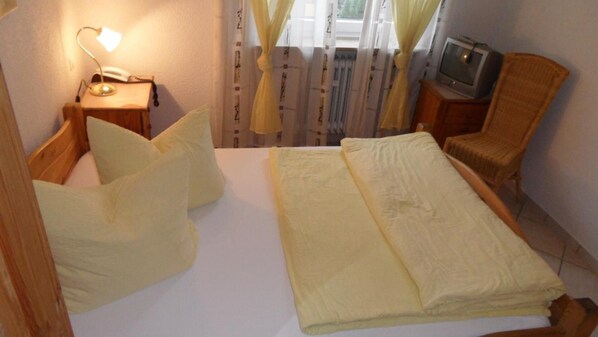 Uma cama de solteiro com colchão branco e duas almofadas, uma luminária de cabeceira, uma mesinha com um telefone, uma televisão sobre um suporte de madeira e uma cadeira.