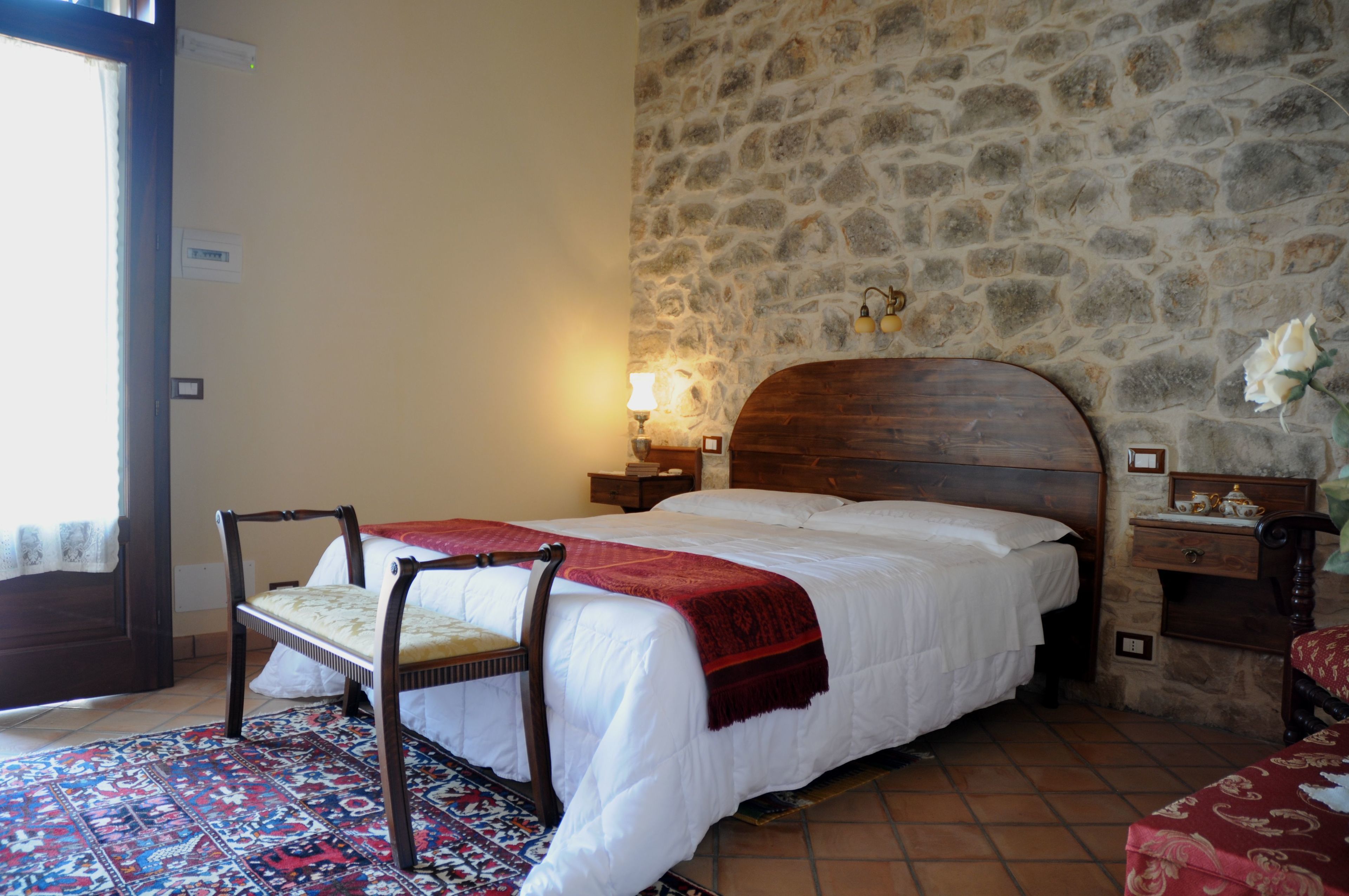 Quarto com parede de pedra, estrutura de cama de madeira, mesa de cabeceira com abajur, uma cadeira e tapete estampado.