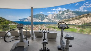 Espaço de exercícios ao ar livre com bicicletas ergométricas e assentos, com vista para uma paisagem montanhosa e um corpo de água.