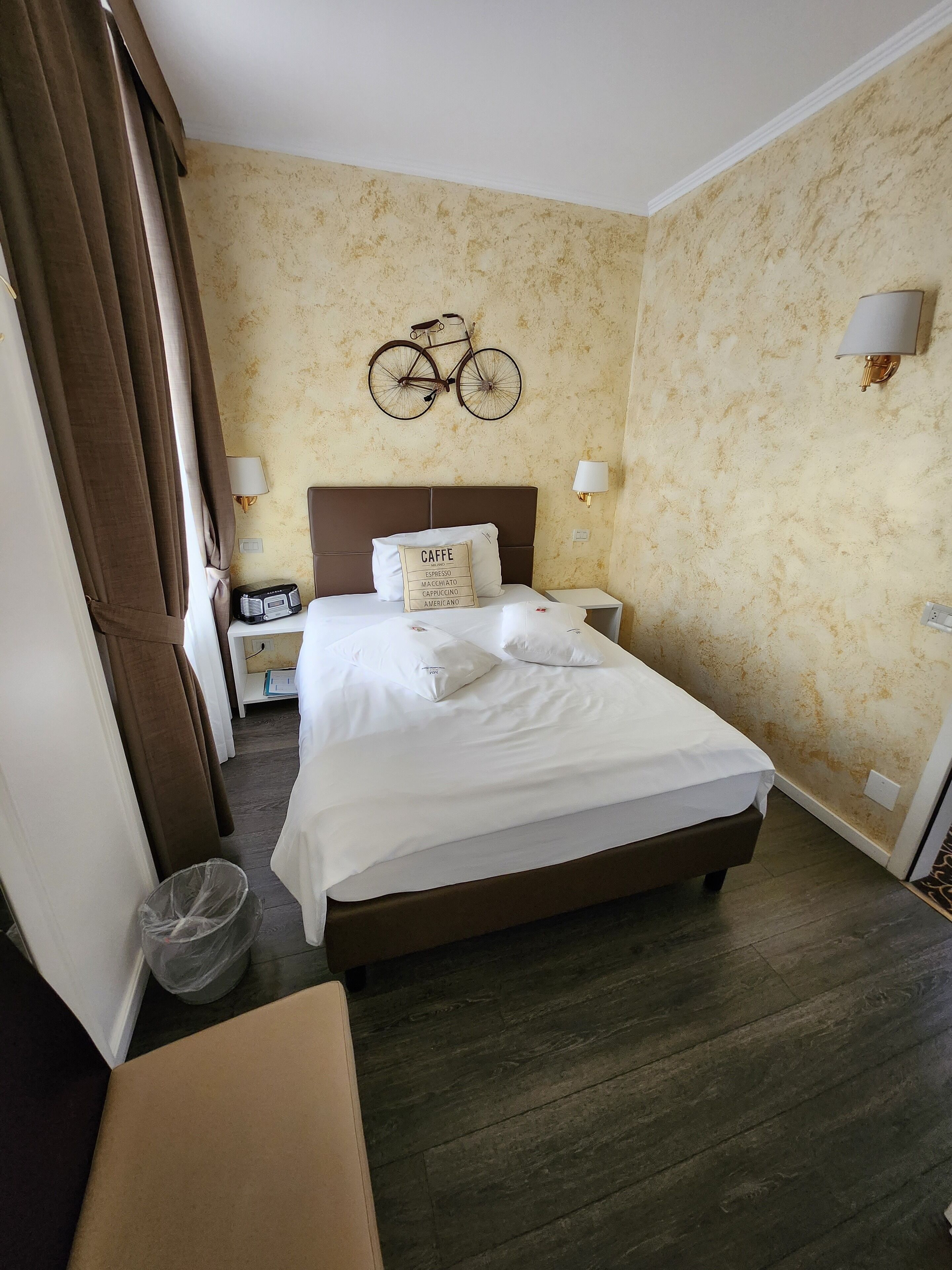 Quarto de hotel com cama, mesa de cabeceira, abajur e bicicleta decorativa na parede.