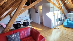 Quarto de sótão com vigas de madeira aparentes, um sofá de couro vermelho com almofada, um sofá azul e uma área de jantar com mesa e cadeiras.