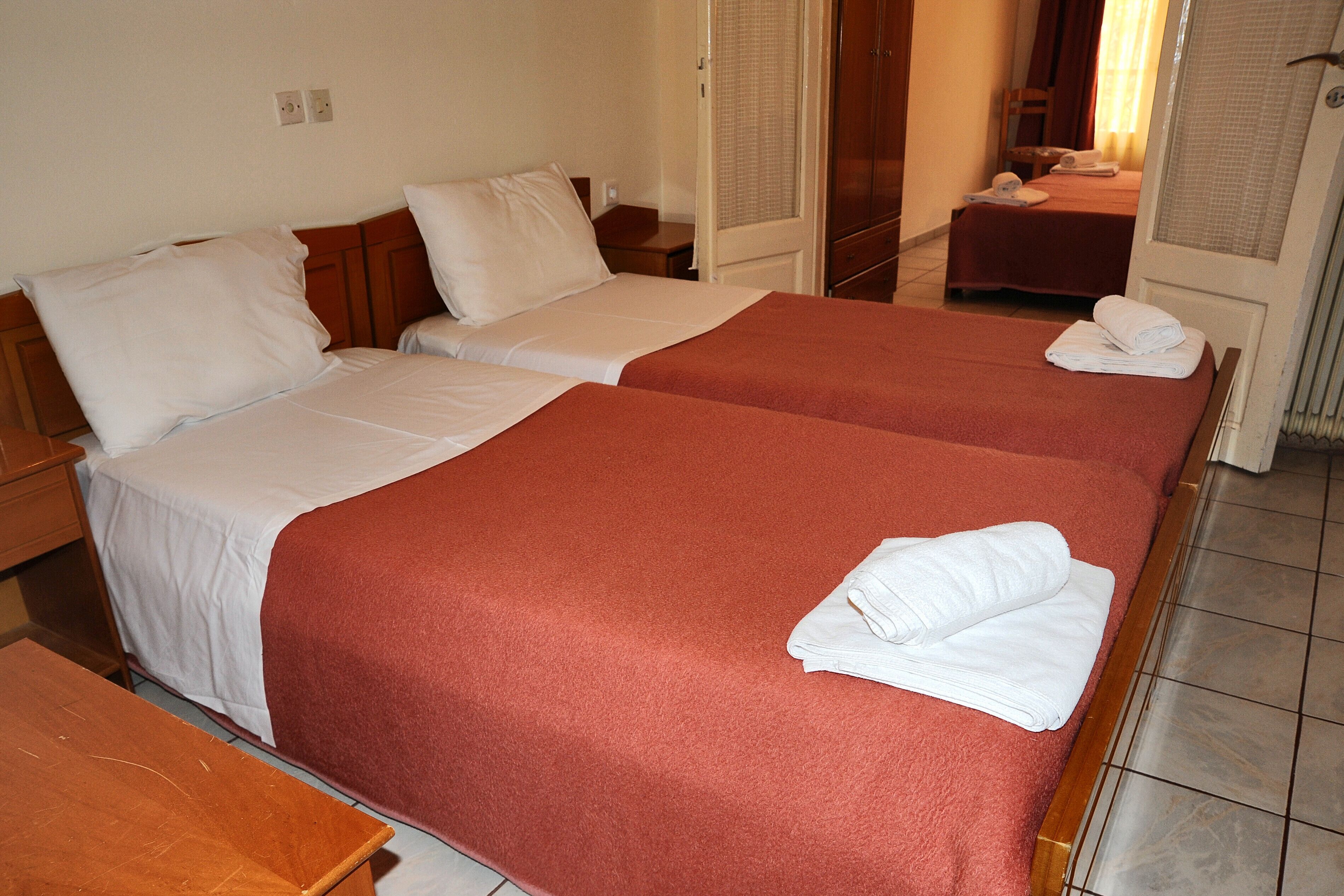 Quarto de hotel com duas camas, cada uma com roupa de cama branca e colchas vermelhas. Há duas mesas de cabeceira, um banco de madeira e um banheiro visível ao fundo.