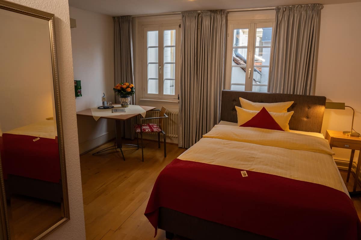 Quarto de hotel com cama, escrivaninha, cadeira e janela com cortinas.