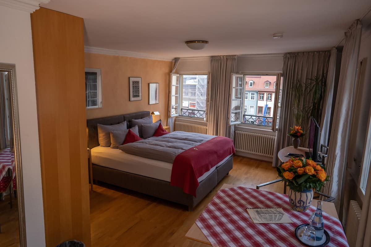 Quarto com cama, janela com vista para edifícios, radiador e mesa com um vaso de flores.