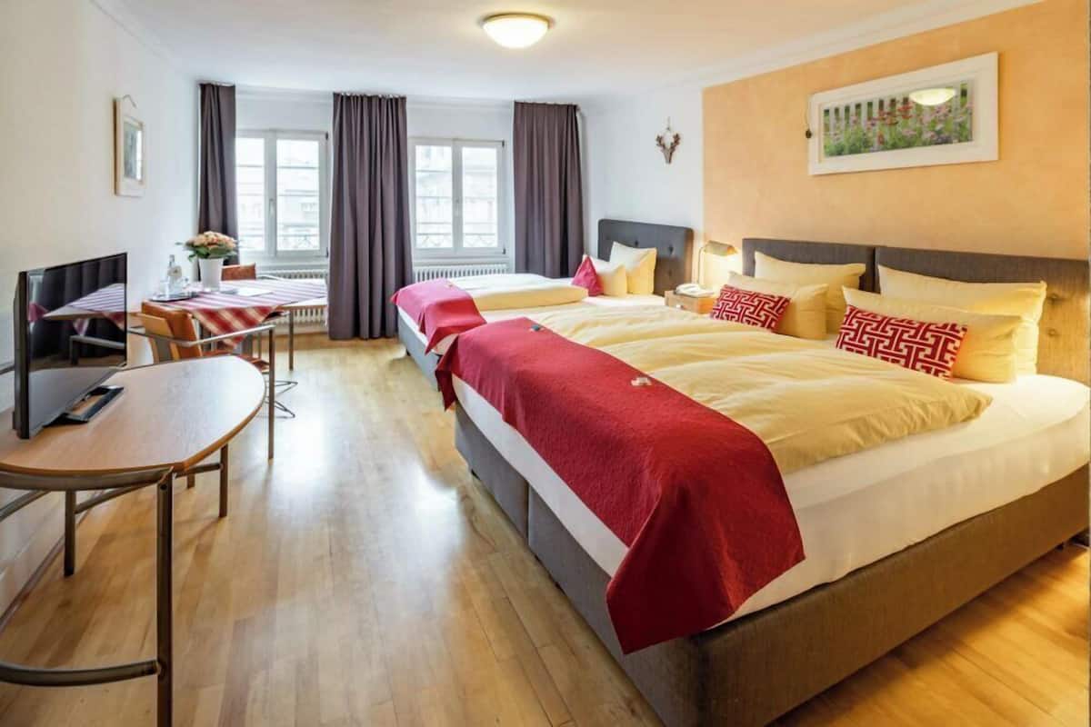 Quarto de hotel com uma cama grande, uma escrivaninha e uma cadeira. Há uma janela com cortinas, um quadro na parede e um abajur na mesa de cabeceira.