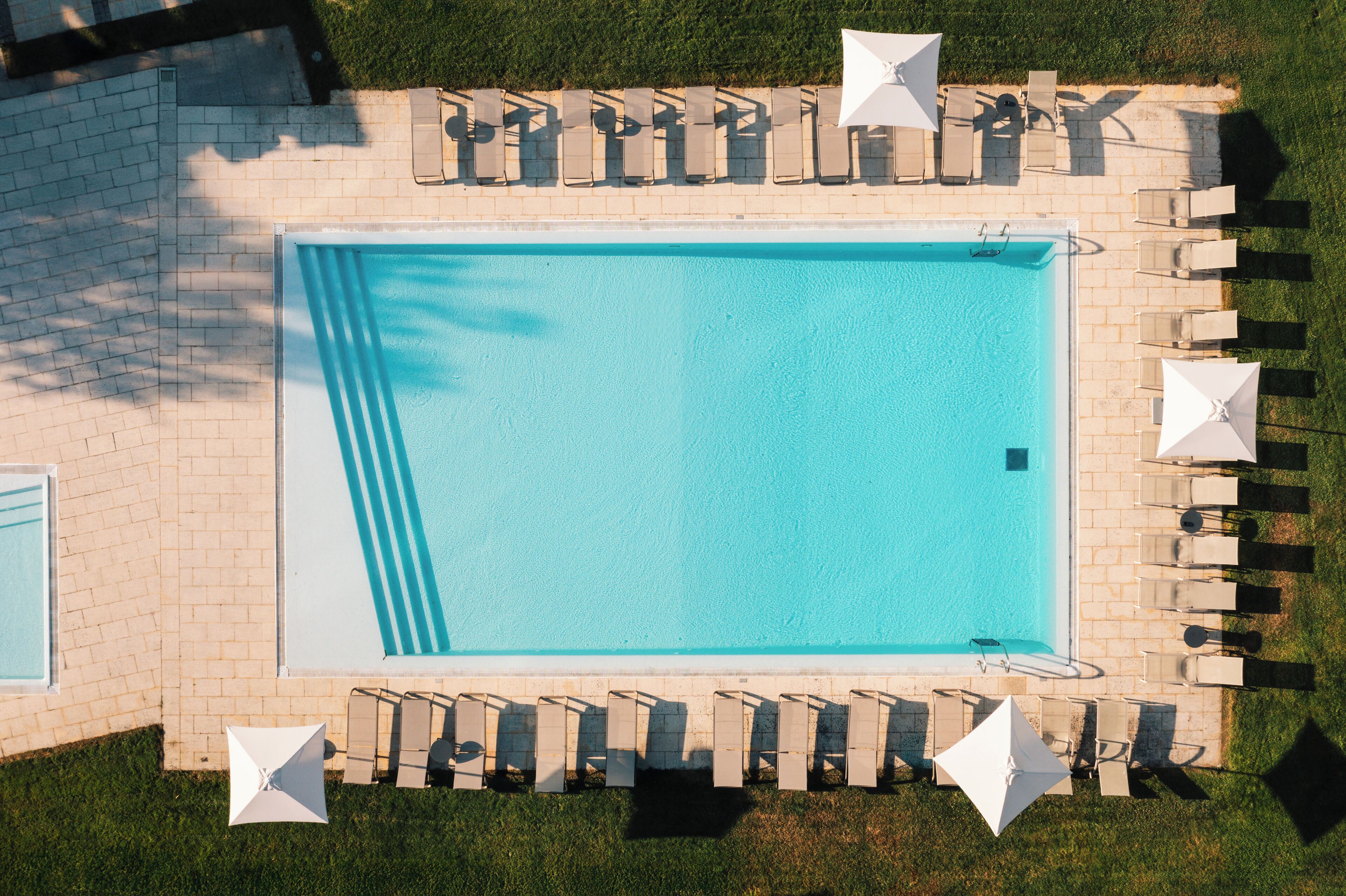 Vista aérea de uma piscina retangular cercada por um pátio de tijolos, com espreguiçadeiras e guarda-sóis dispostos ao redor.